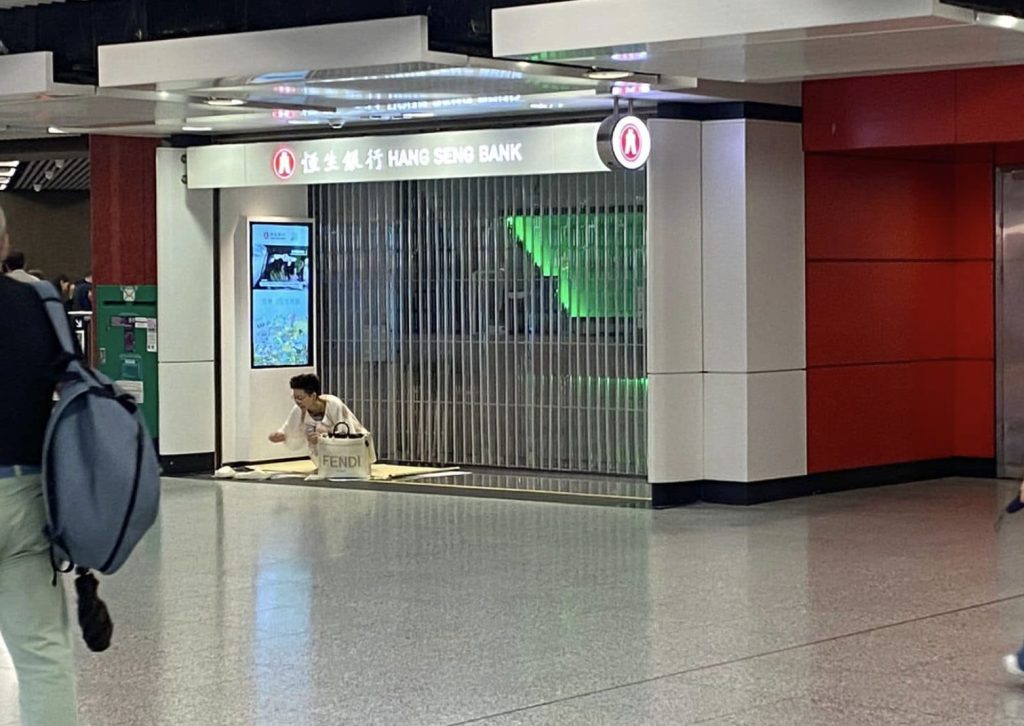 Middle-aged woman lies down in front of Hang Seng Bank in MTR Central Station to read newspapers ...